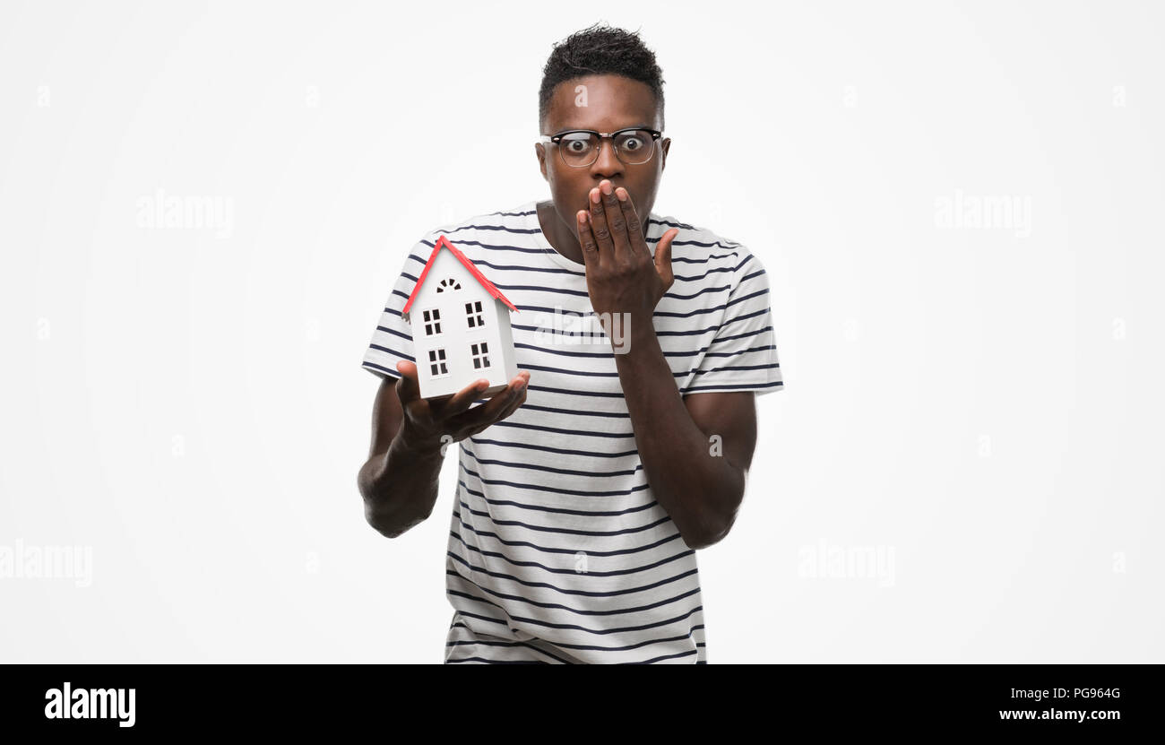 Young african american man holding house cover mouth with hand shocked ...