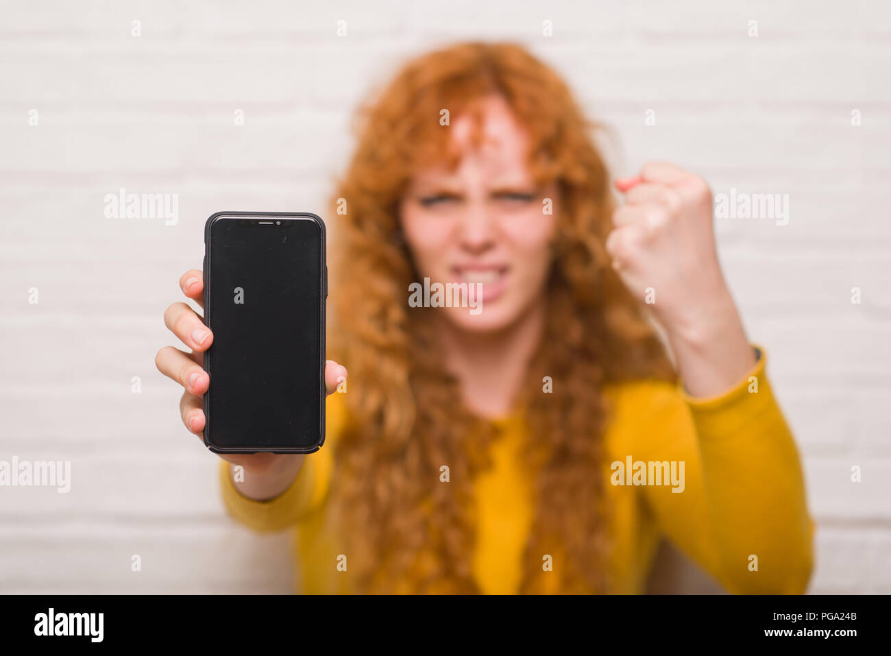 Young redhead woman sitting over brick wall showing smartphone annoyed ...