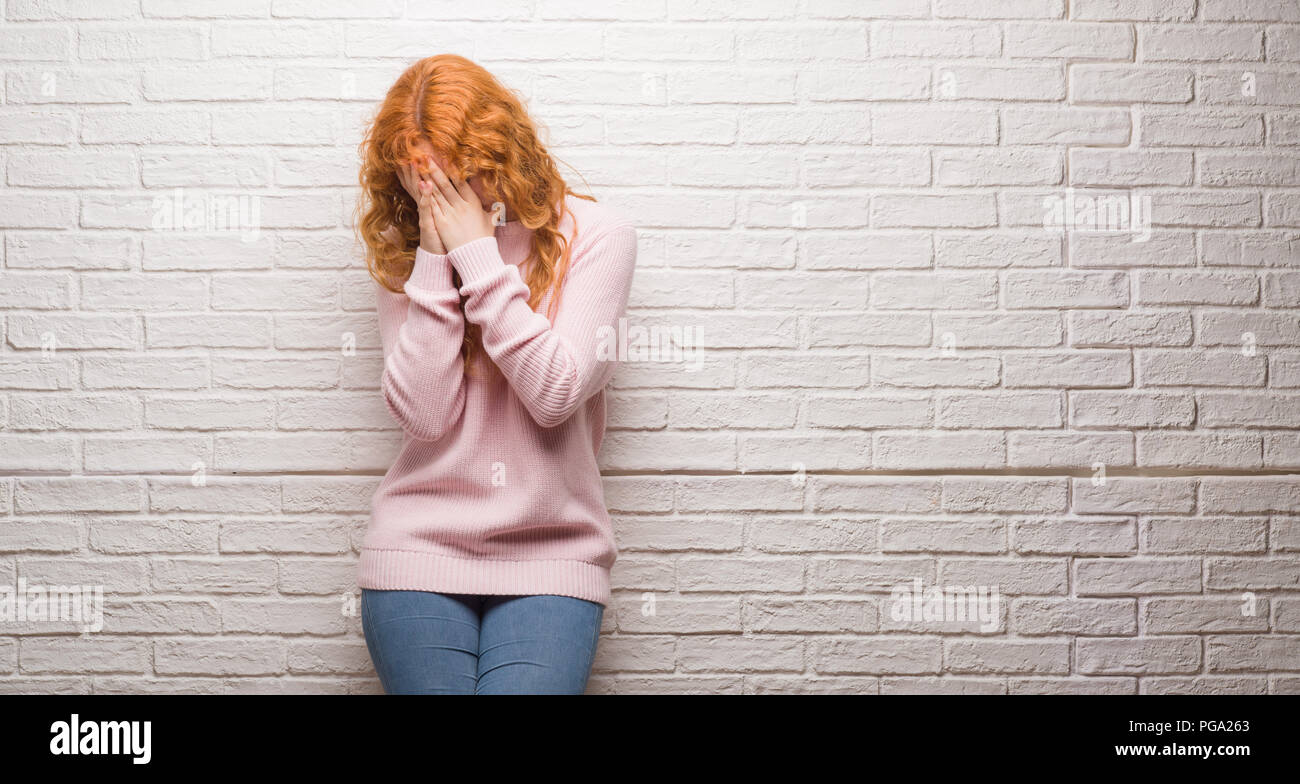 Young redhead woman standing over brick wall with sad expression ...