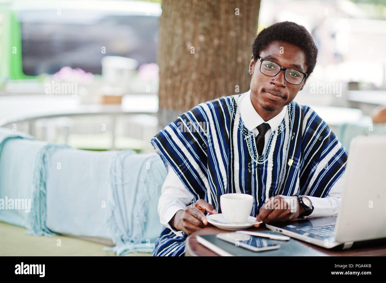 African man in traditional clothes and glasses sitting at outdoor caffe ...