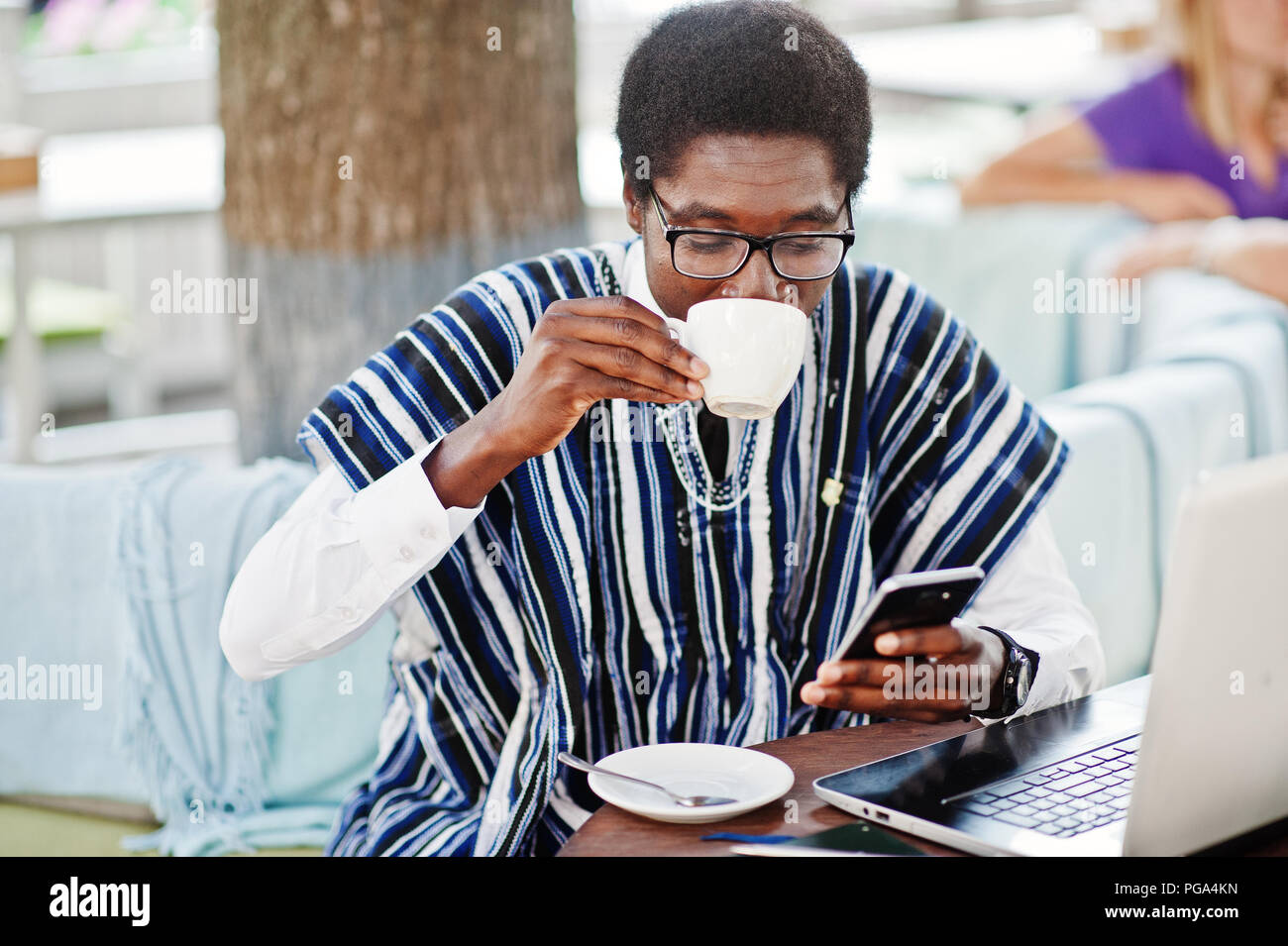 African man in traditional clothes and glasses sitting at outdoor caffe ...