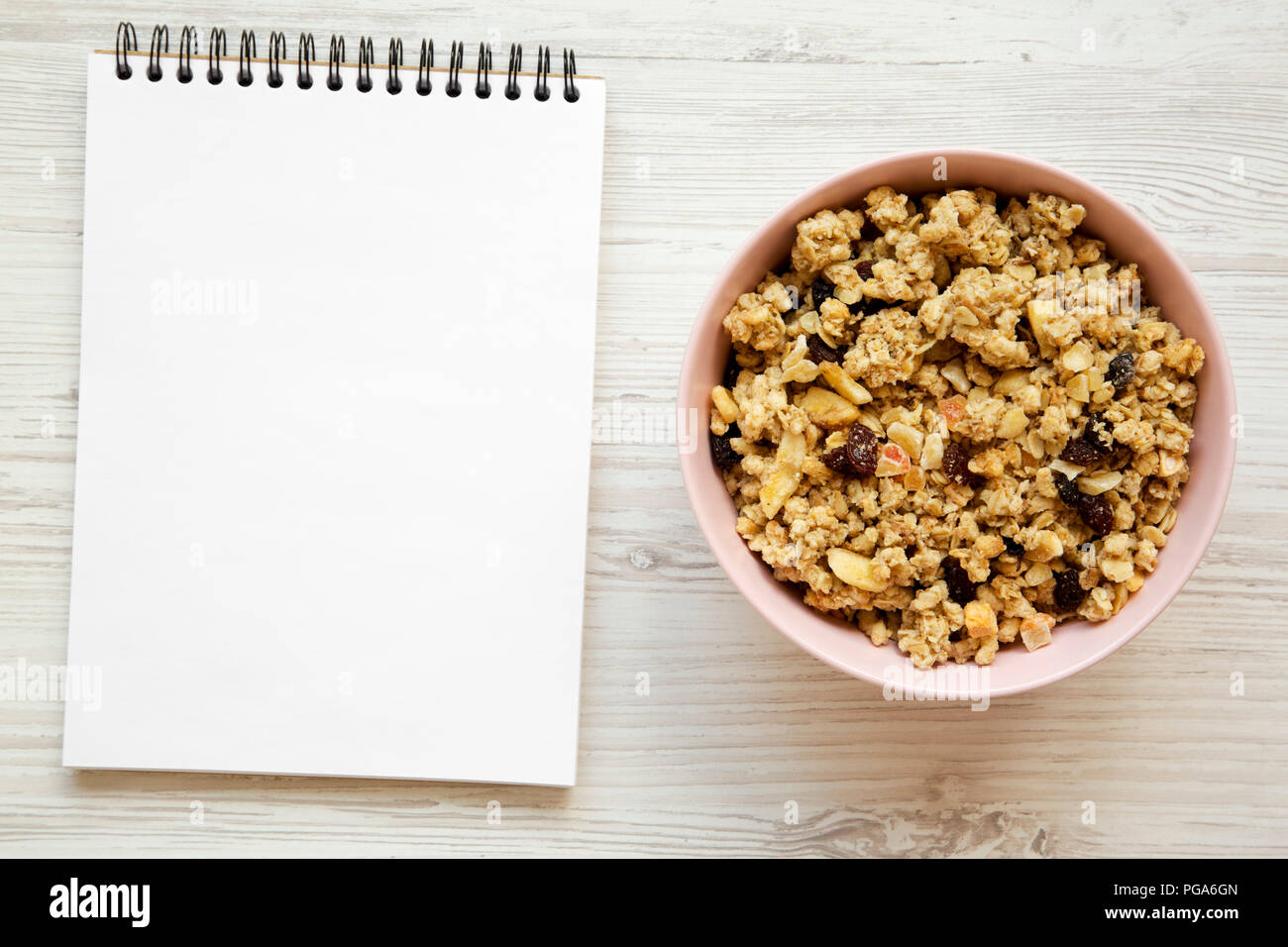 Pink bowl of fruit granola and blank notepad on white wooden background ...