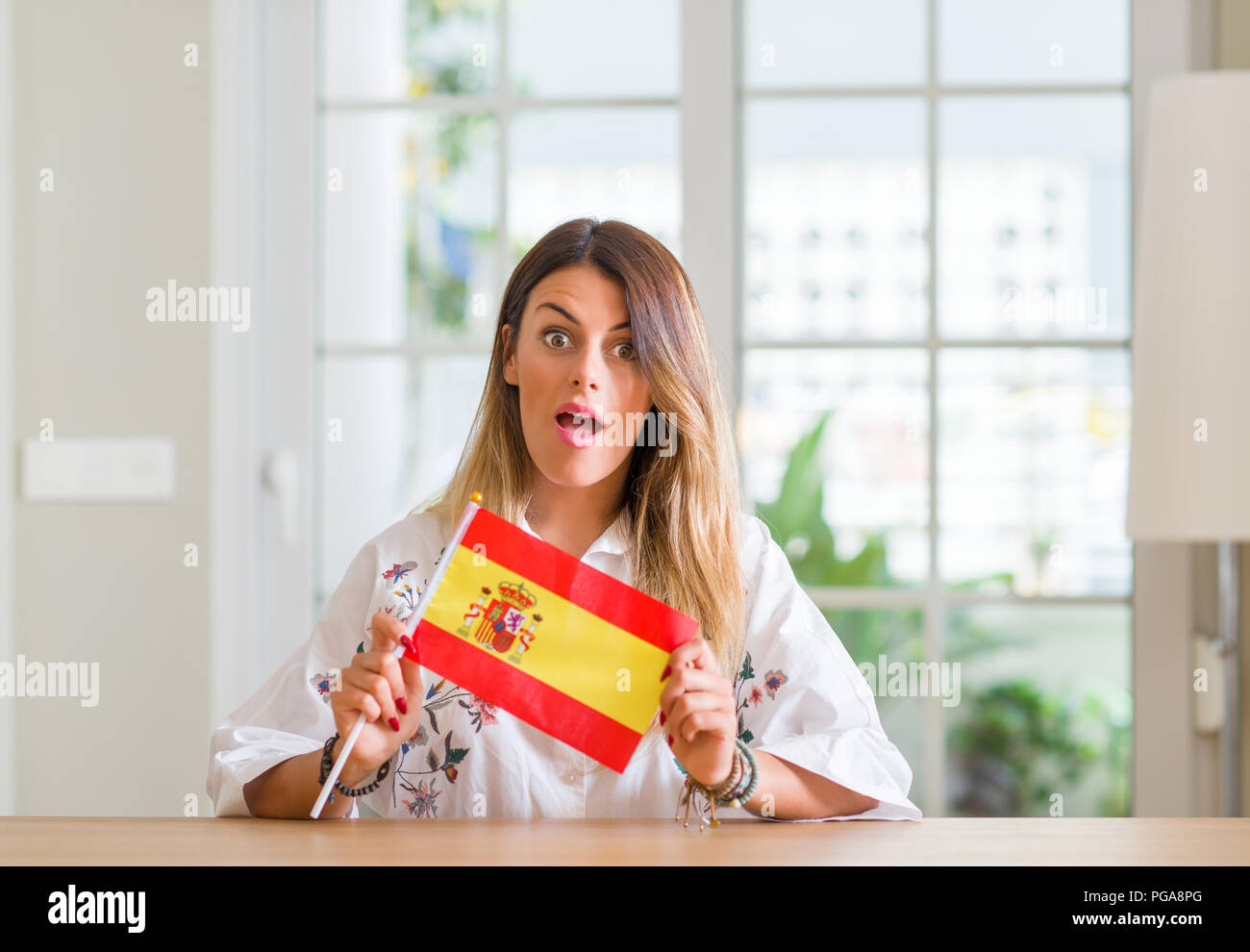 Young woman at home holding flag of Spain scared in shock with a ...