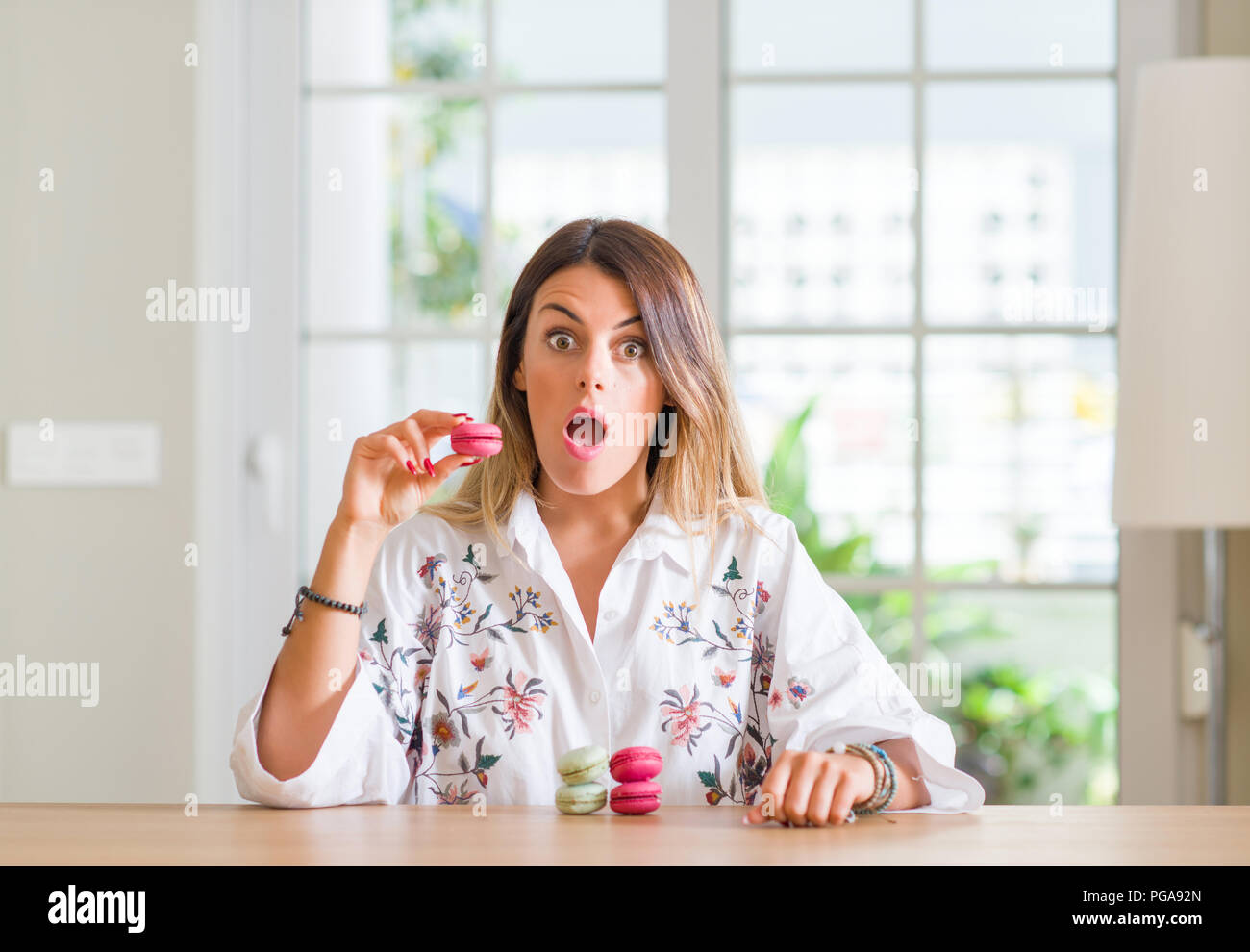 Young woman at home eating macarons scared in shock with a surprise ...