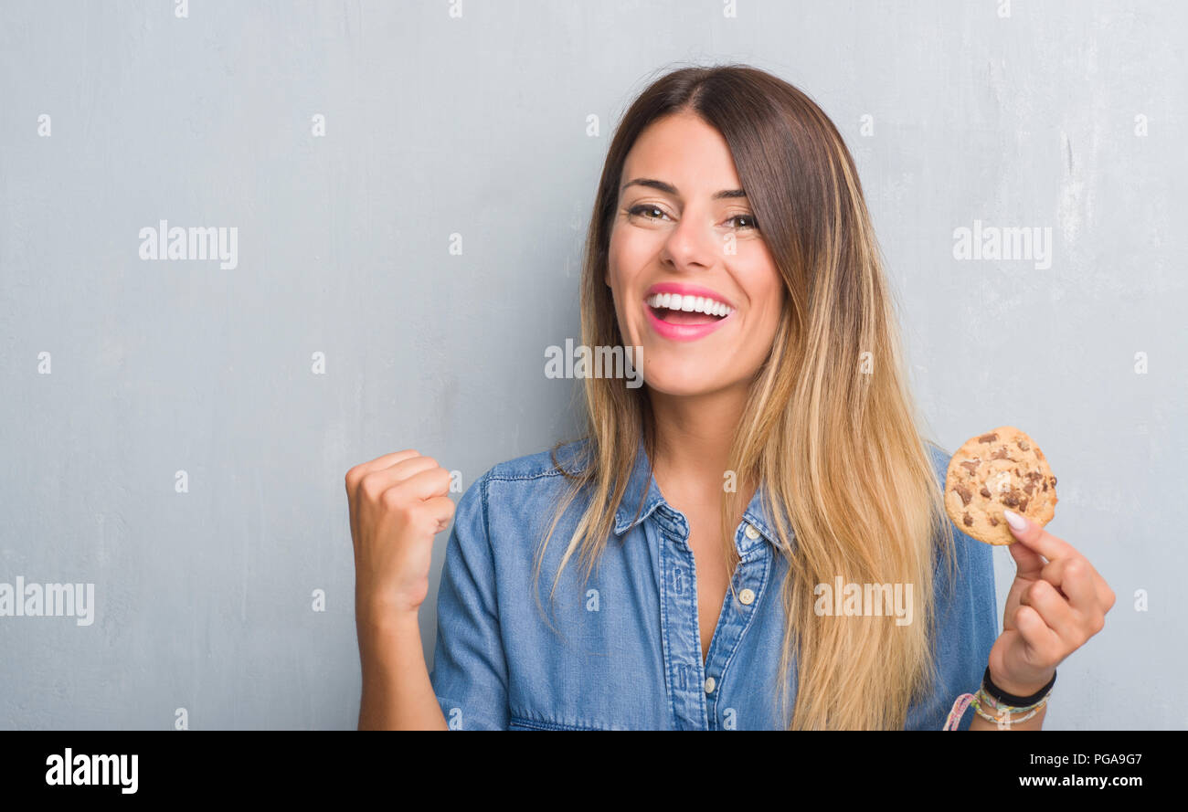 Young adult woman over grey grunge wall eating chocolate chip cooky ...