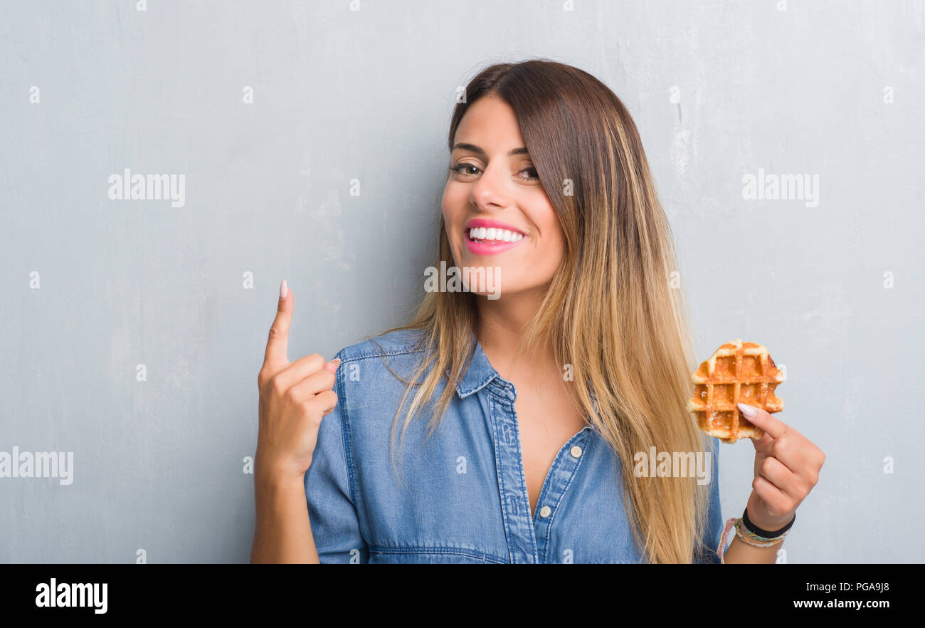 Young adult woman over grey grunge wall eating Belgium waffle surprised ...