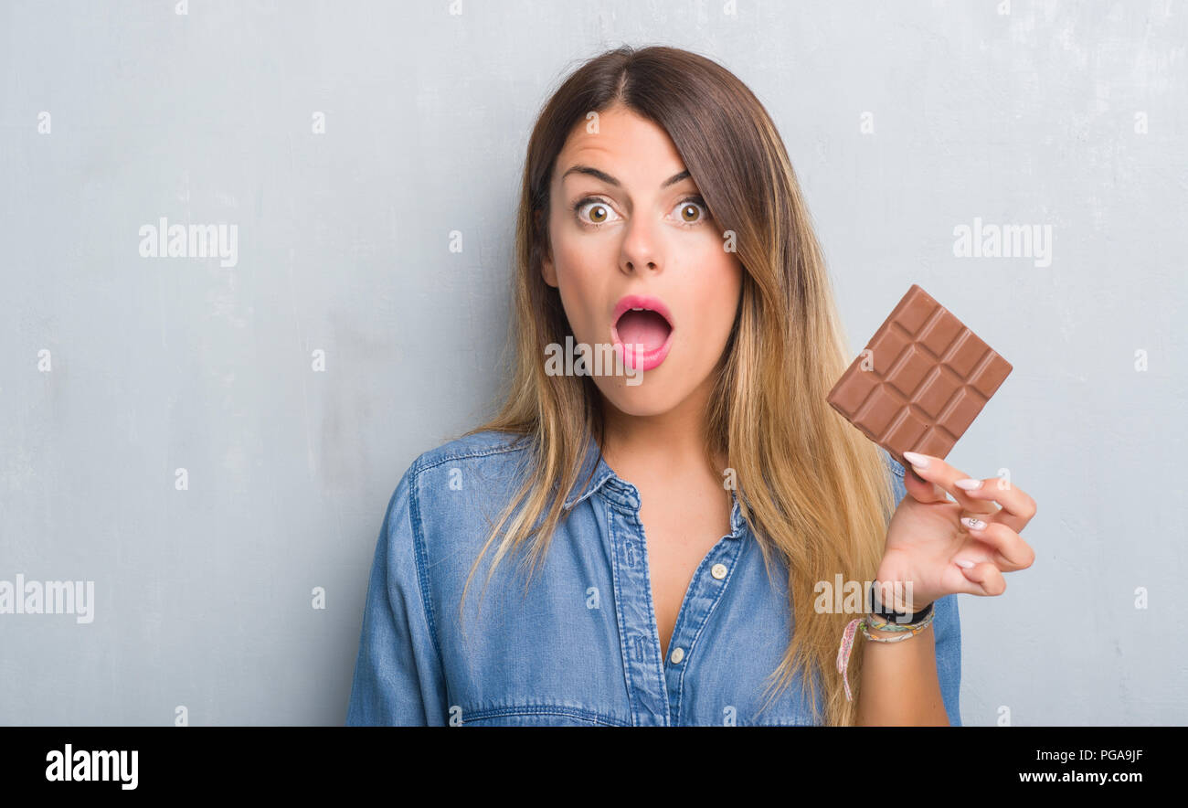 Young adult woman over grey grunge wall eating chocolate bar scared in ...