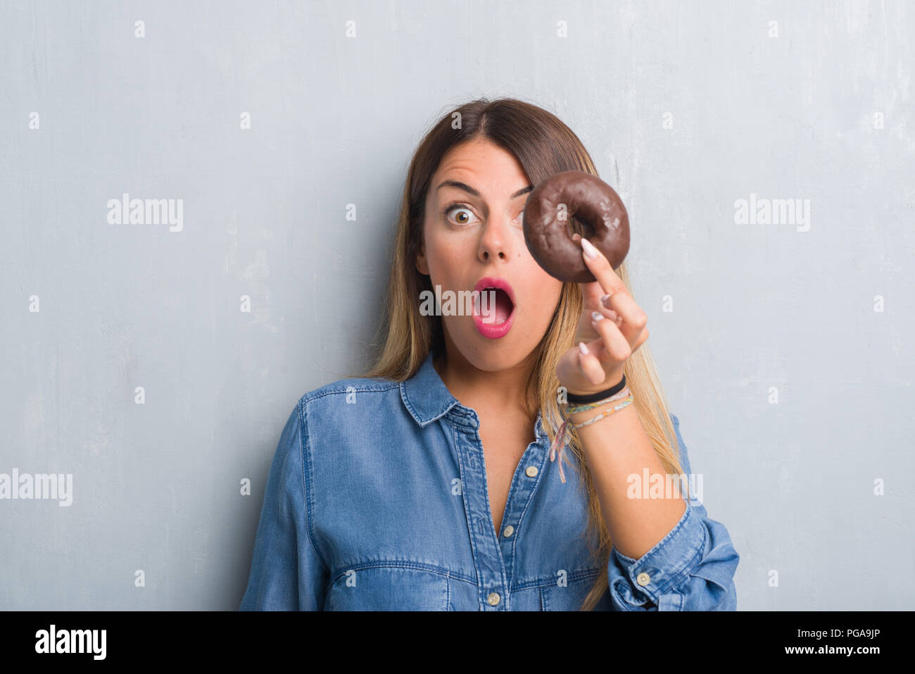 Young adult woman over grey grunge wall eating chocolate donut scared ...