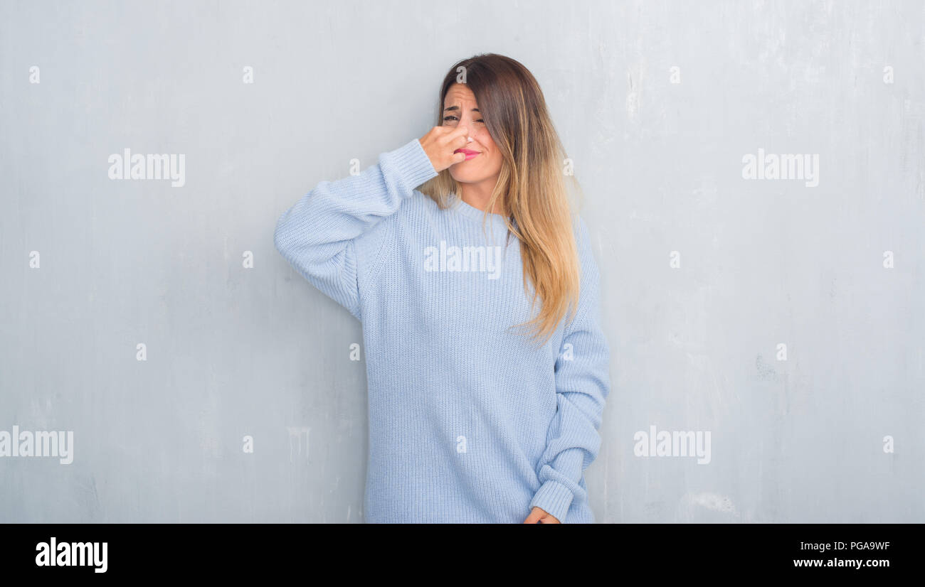 Young adult woman over grey grunge wall wearing winter outfit smelling ...