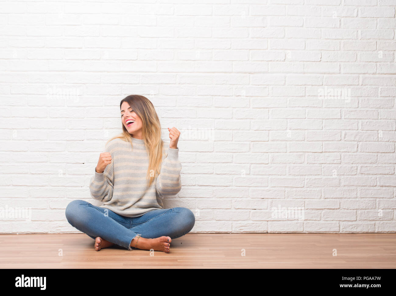Young adult woman sitting on the floor over white brick wall at home ...