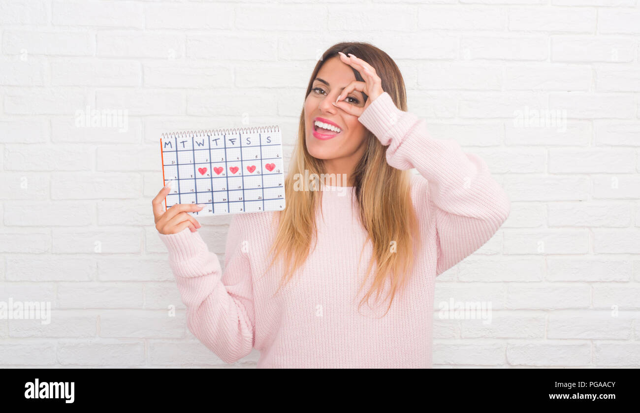 Young adult woman over white brick wall holding period calendar with ...