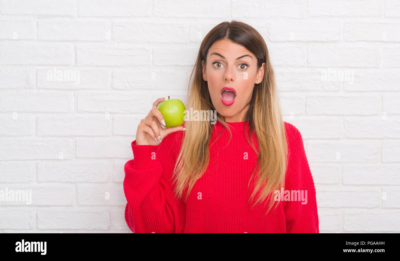 Young adult woman over white brick wall eating fresh green apple scared ...