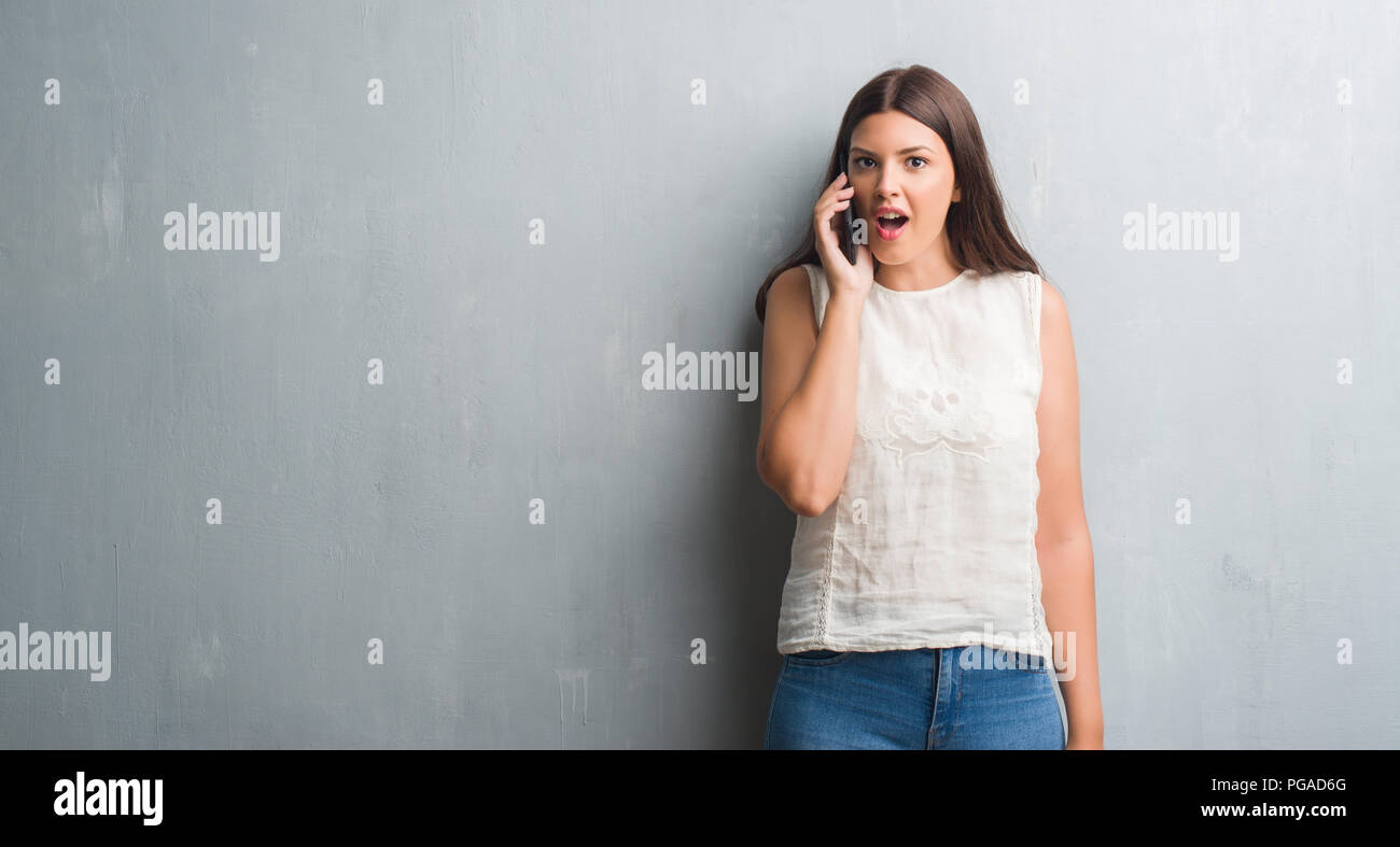 Young brunette woman over grunge grey wall talking on the phone scared ...