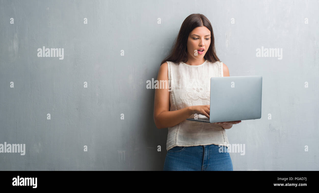 Young brunette woman over grunge grey wall using computer laptop scared ...