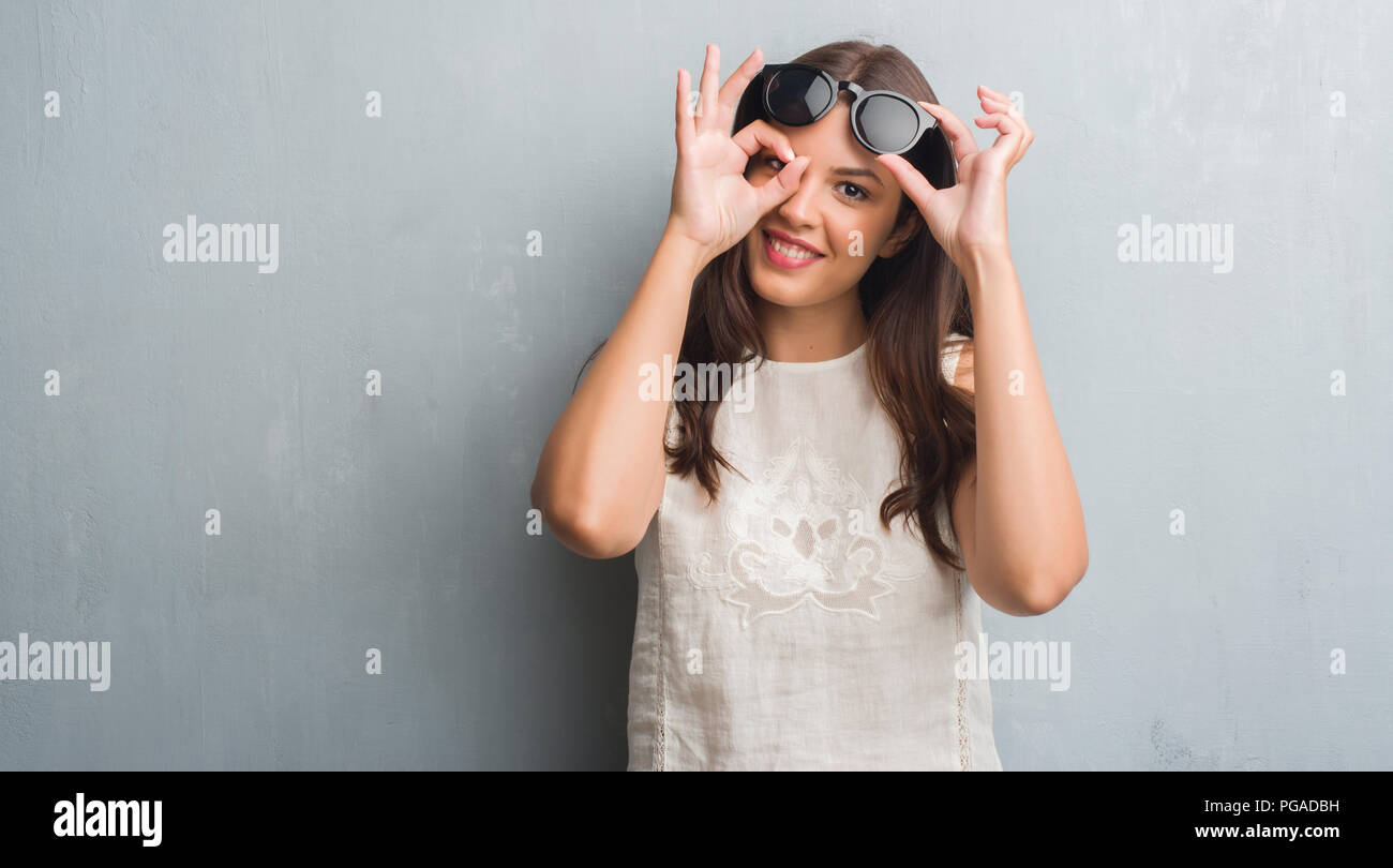 Young brunette woman over grunge grey wall wearing fashion sunglasses ...