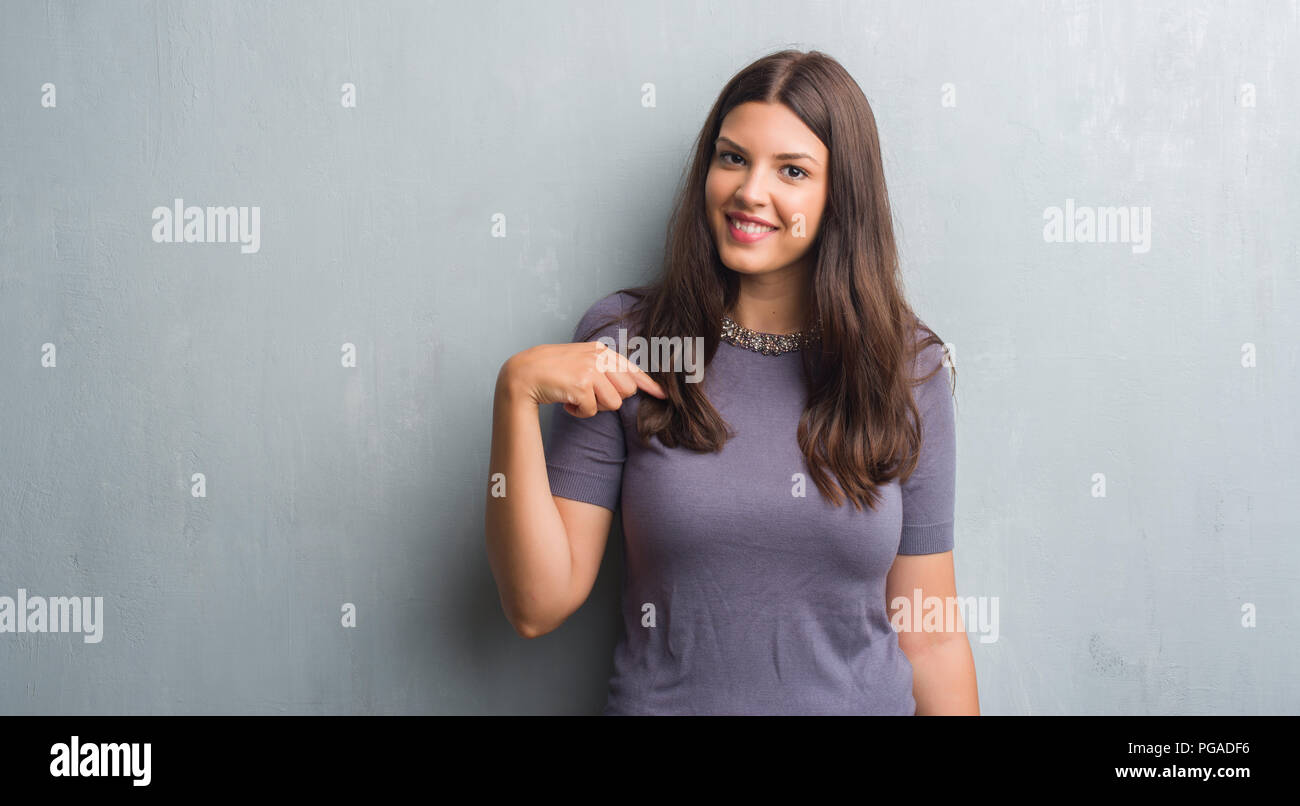 Young brunette woman over grunge grey wall with surprise face pointing ...