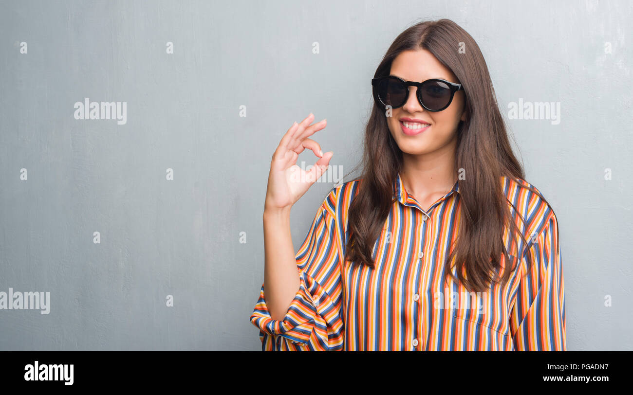 Young brunette woman over grunge grey wall wearing big sunglasses doing ...
