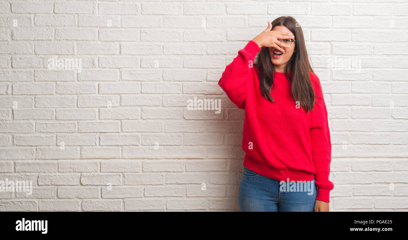 Young brunette woman standing over white brick wall peeking in shock ...