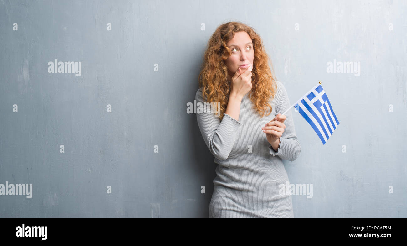Young redhead woman over grey grunge wall holding flag of Greece ...