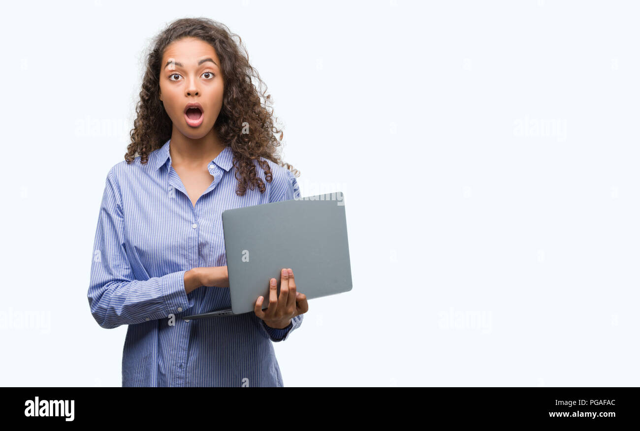 Young hispanic woman holding computer laptop scared in shock with a ...