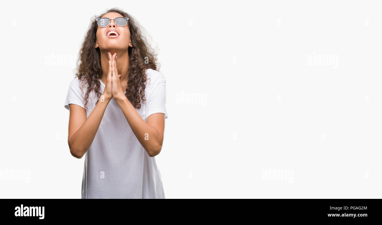 Young hispanic woman wearing sunglasses begging and praying with hands ...