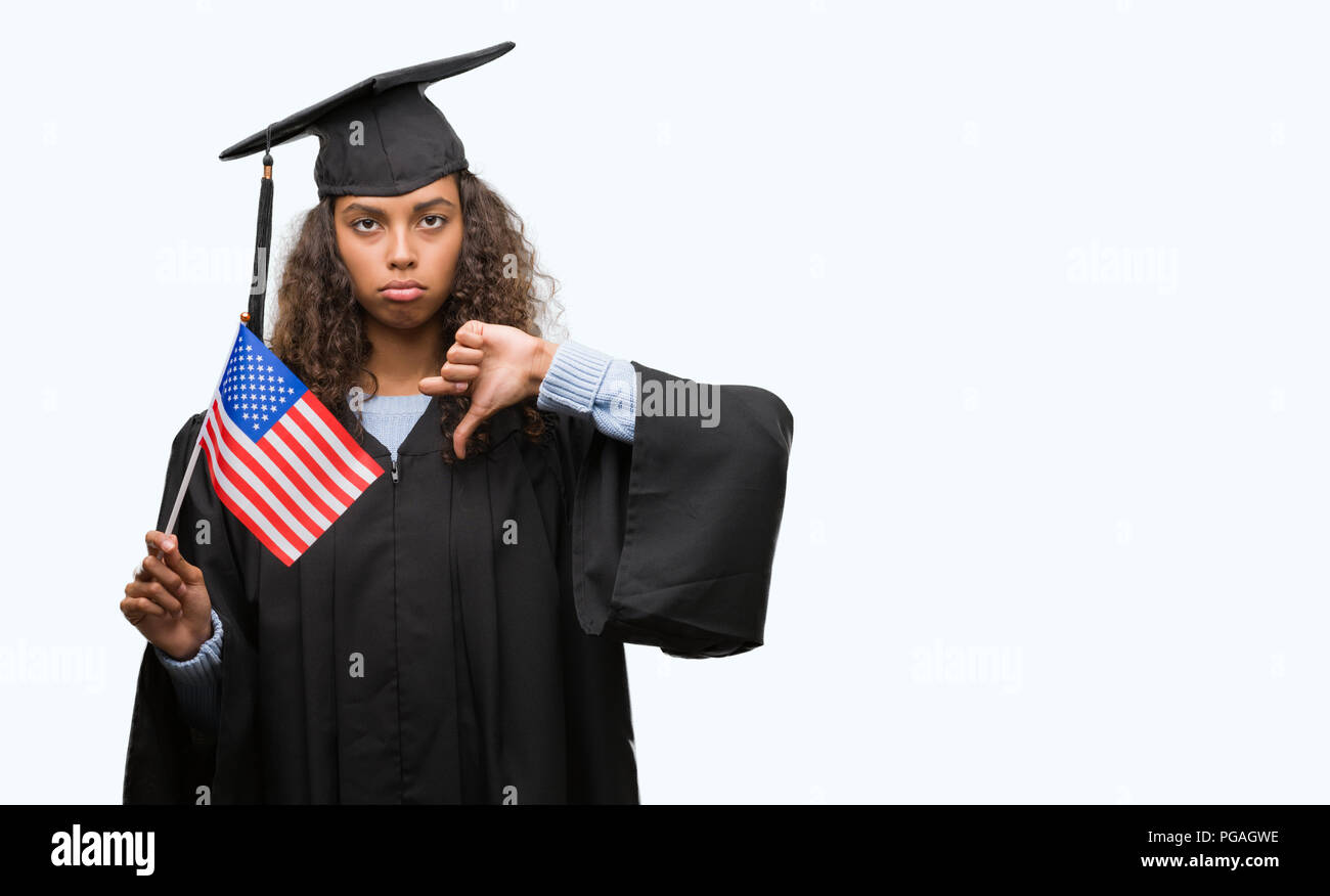 Young hispanic woman wearing graduation uniform holding flag of United ...