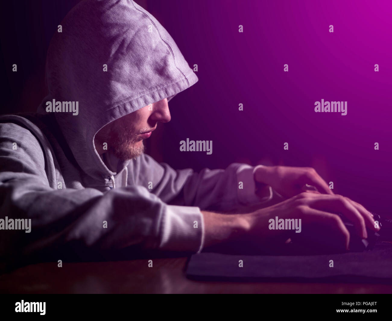 portrait of hooded man typing text on keyboard in front of pc monitor ...