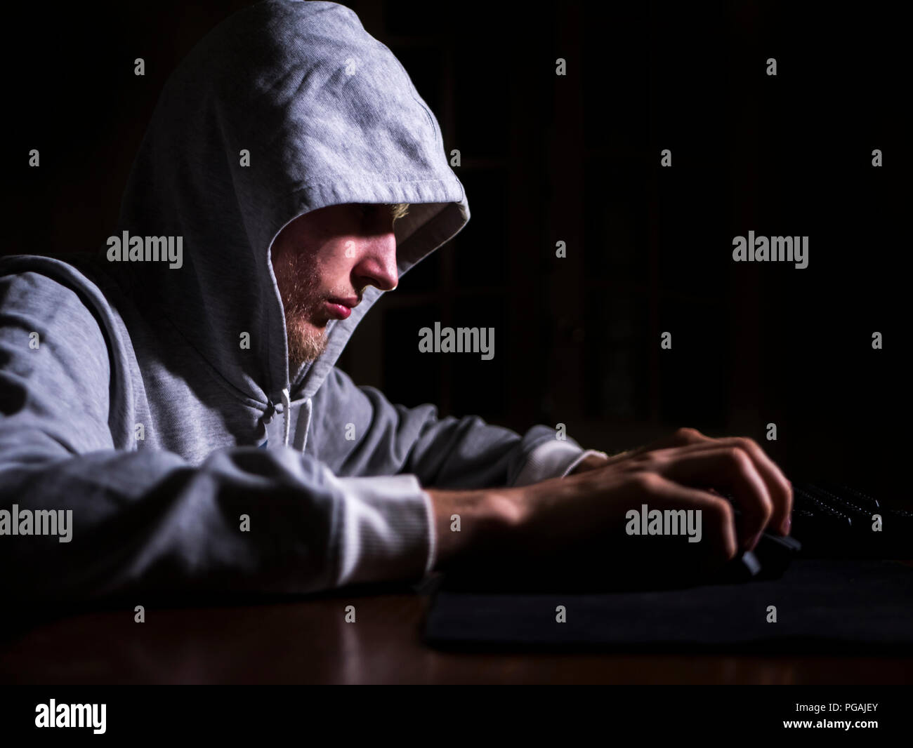 portrait of hooded man typing text on keyboard in front of pc monitor ...