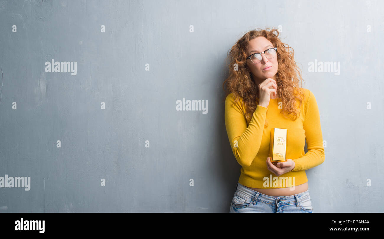 Young redhead woman over grey grunge wall holding gold ingot serious ...