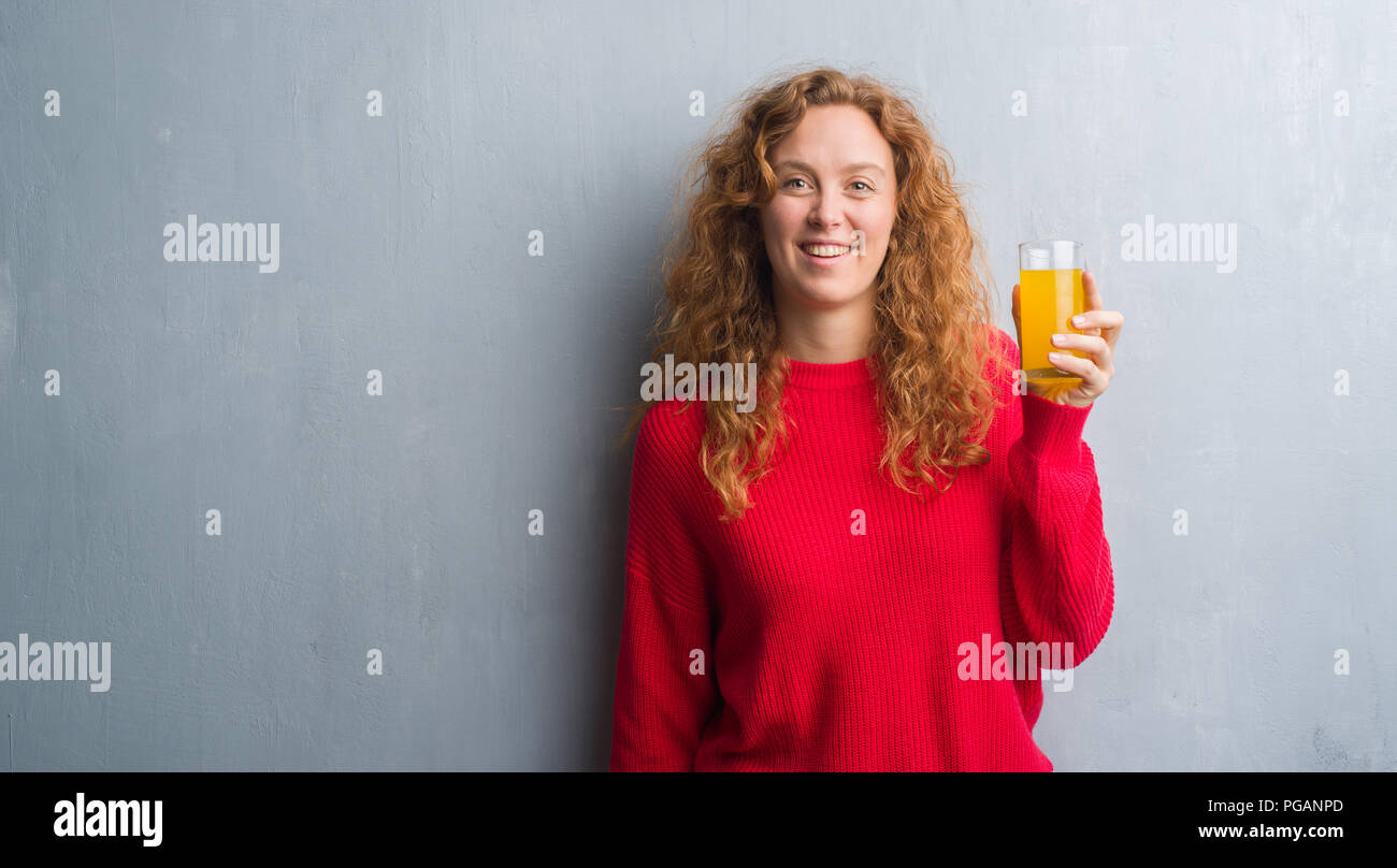Young redhead woman over grey grunge wall drinking a glass of orange ...