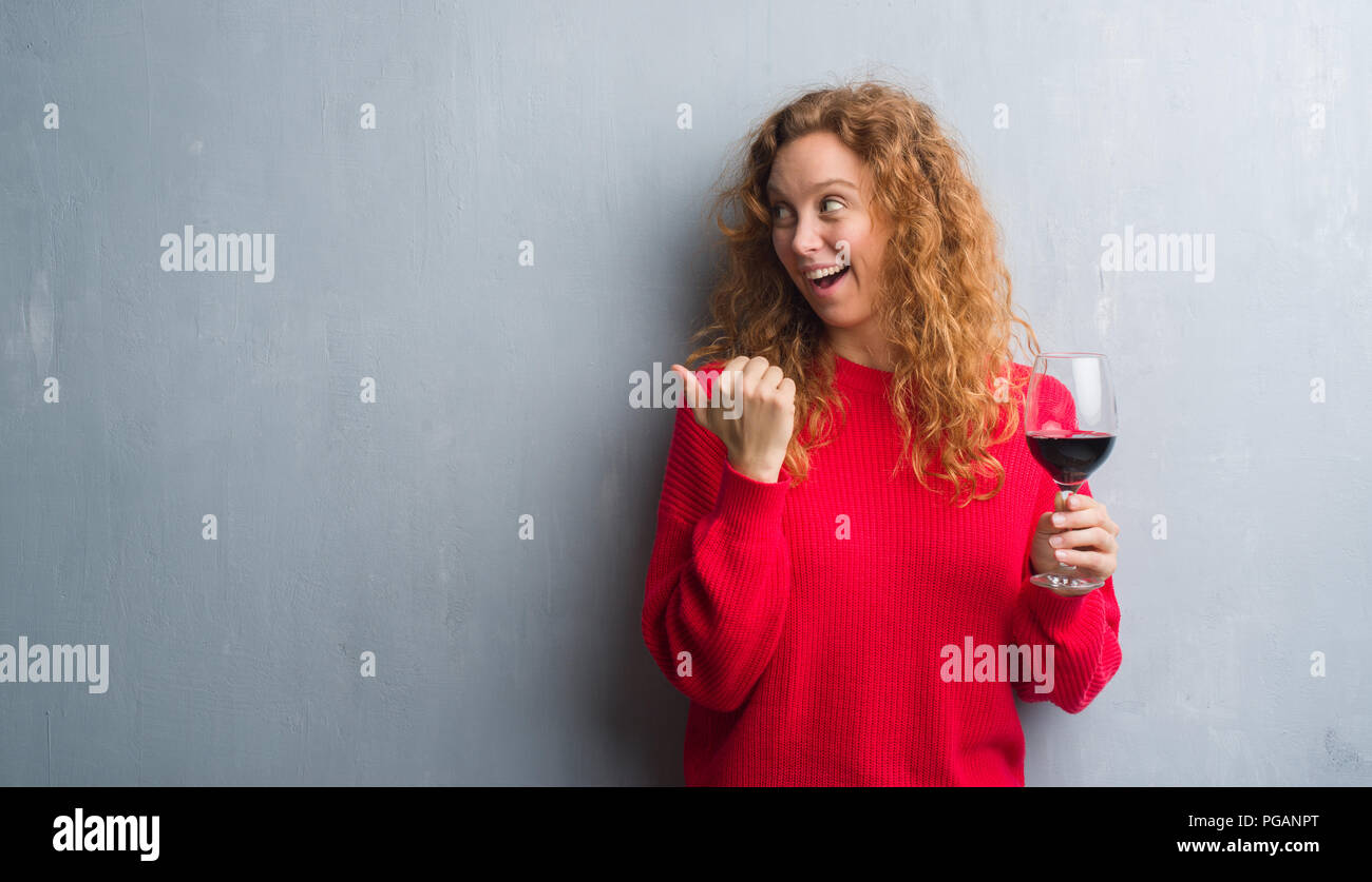 Young redhead woman over grey grunge wall drinking a glass of wine ...