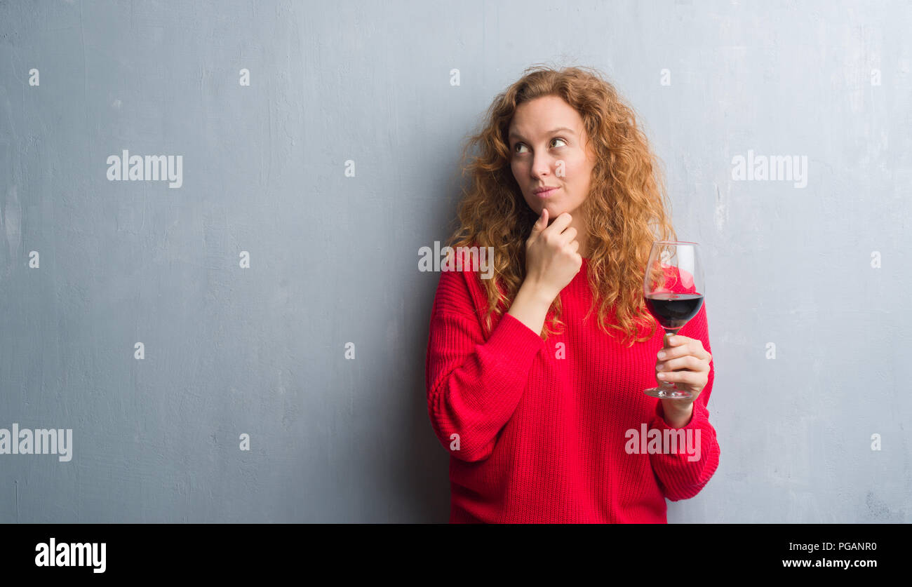 Young redhead woman over grey grunge wall drinking a glass of wine ...