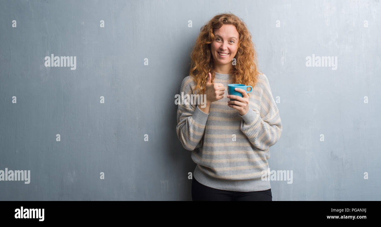 Young redhead woman over grey grunge wall drinking a cup of coffee ...
