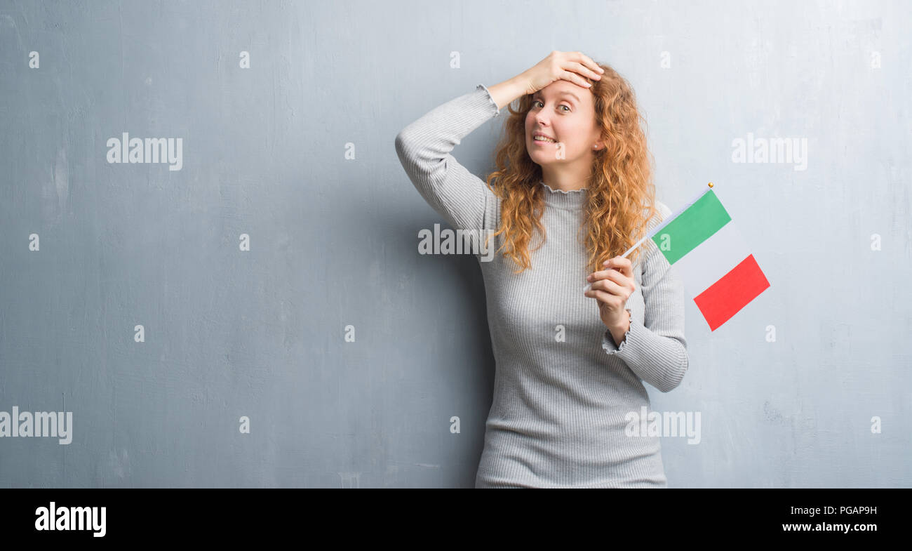 Young redhead woman over grey grunge wall holding flag of Italy ...