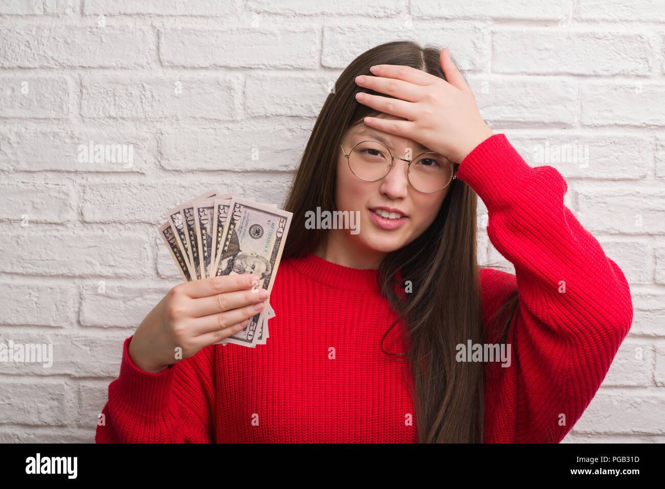 Young Chinese woman over brick wall holding dollars stressed with hand ...
