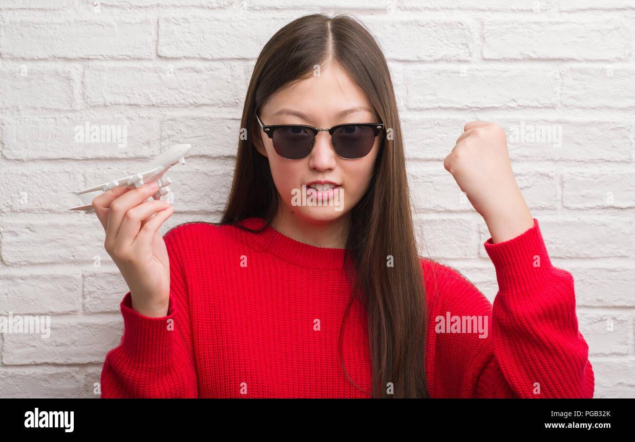 Young Chinese woman over brick wall holding plane toy annoyed and ...
