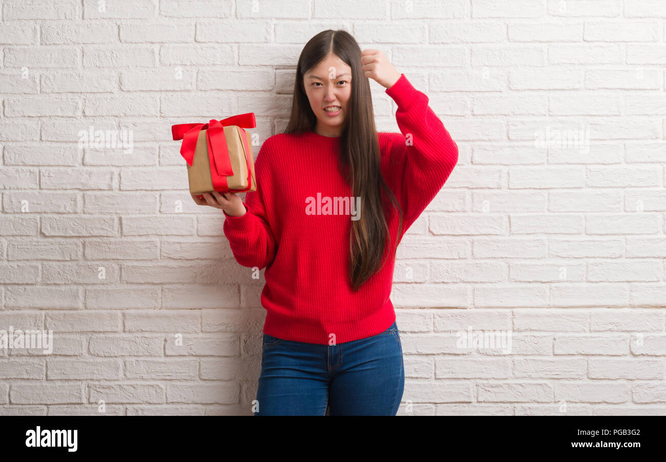 Young Chinese woman over brick wall holding a gift annoyed and ...