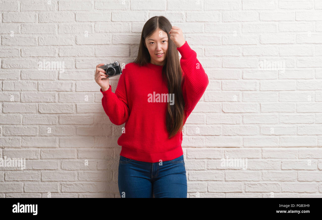 Young Chinese woman over brick wall holding vintage camera annoyed and ...