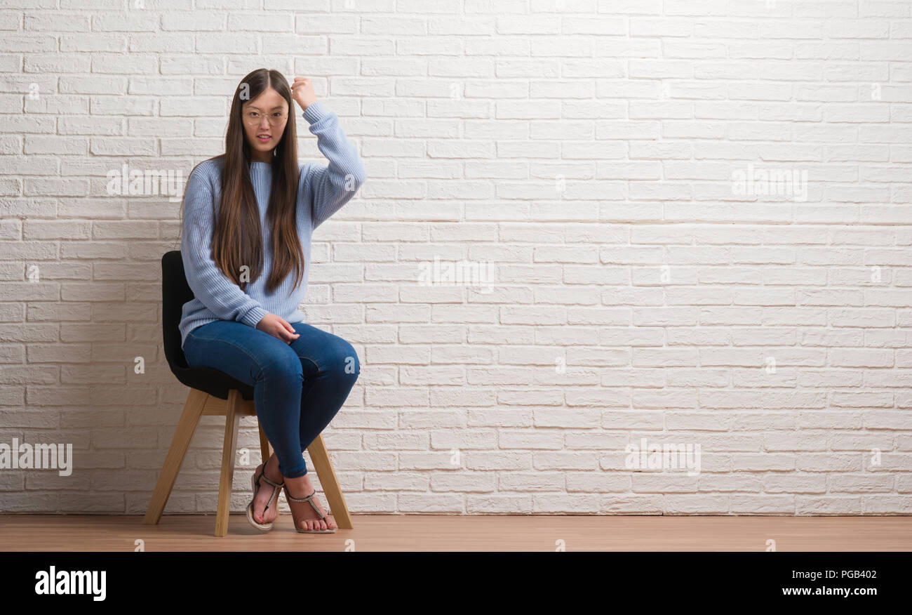 Young Chinese woman sitting on chair over brick wall annoyed and ...