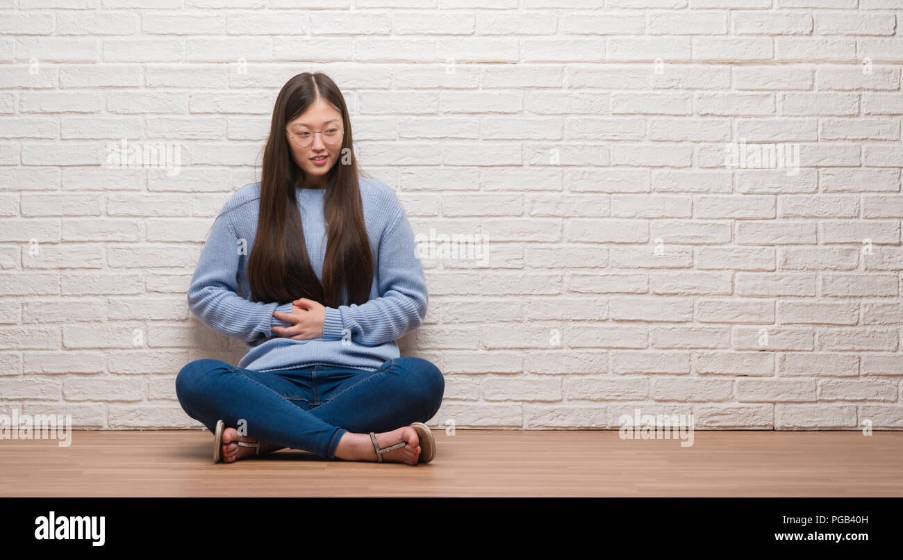 Young Chinese woman sitting on the floor over brick wall with hand on ...