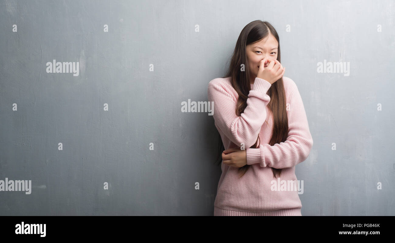 Young Chinese woman over grey wall smelling something stinky and ...