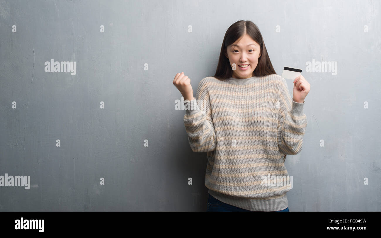 Young Chinese woman over grey wall holding credit card screaming proud ...