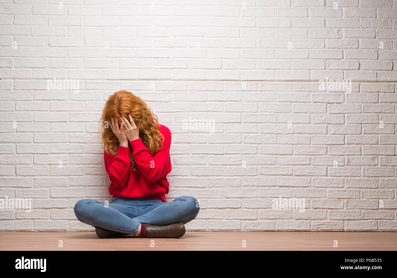 Young redhead woman sitting over brick wall with sad expression ...