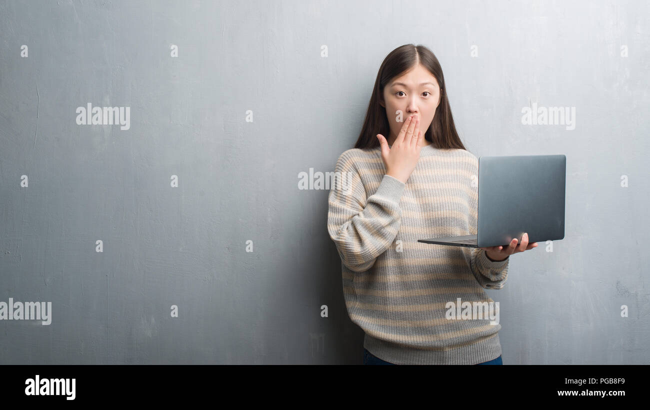 Young Chinese woman over grey wall using computer laptop cover mouth ...