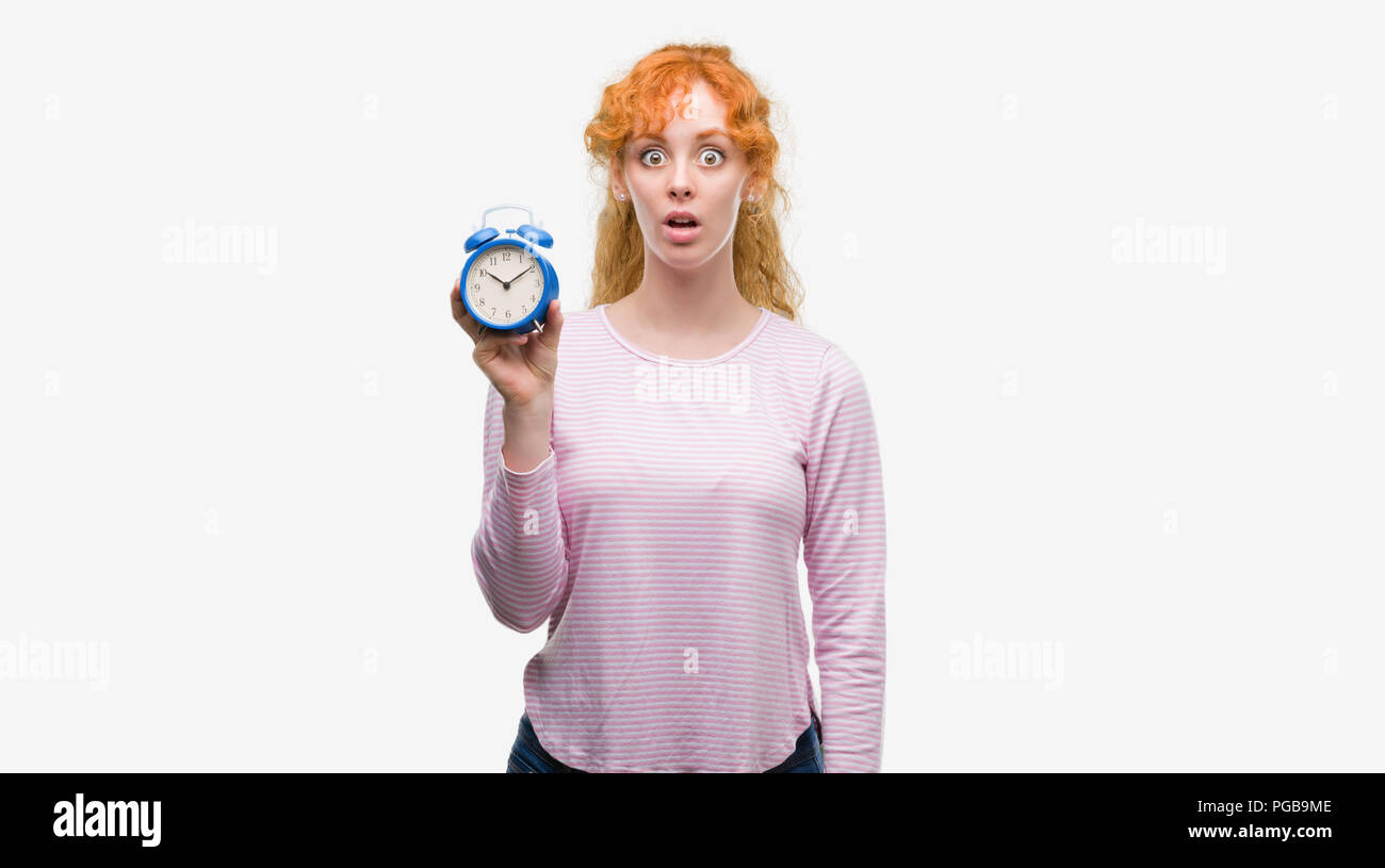 Young redhead woman holding alarm clock scared in shock with a surprise ...