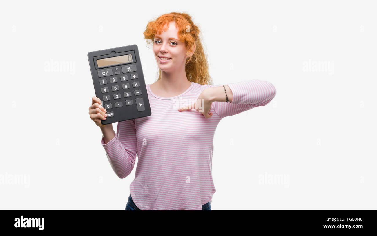 Young redhead woman holding big calculator with surprise face pointing ...