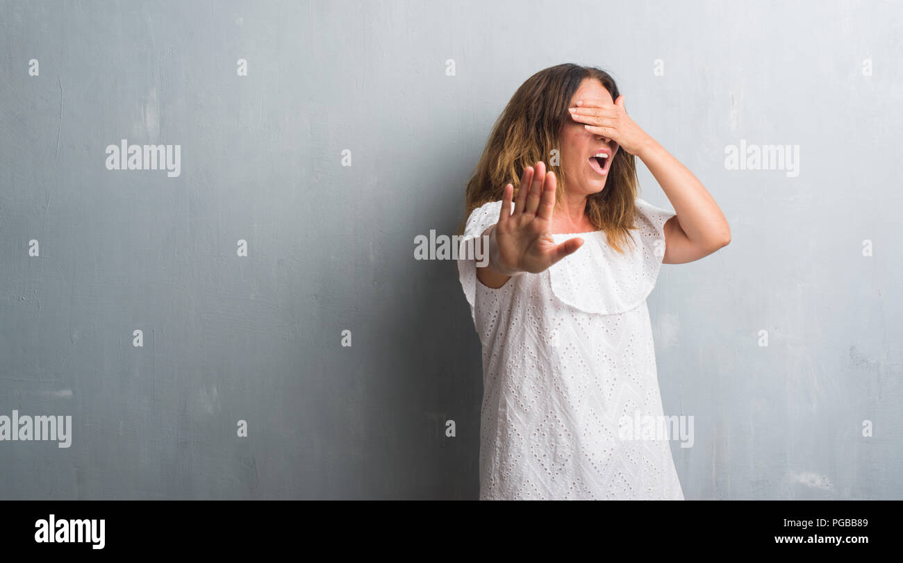 Middle age hispanic woman standing over grey grunge wall covering eyes ...
