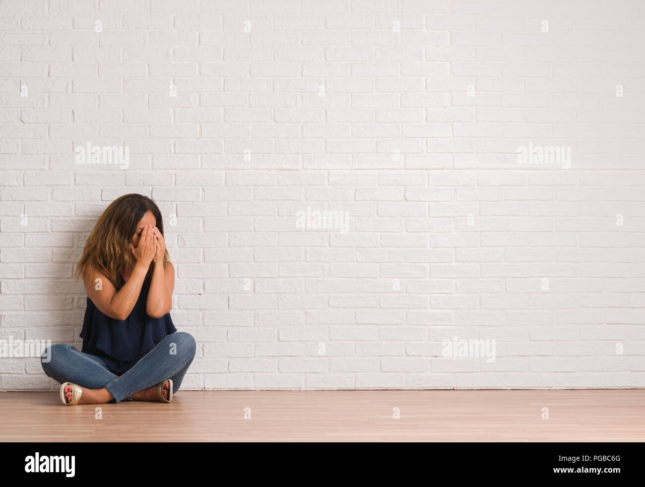 Middle age hispanic woman sitting on the floor over white brick wall ...