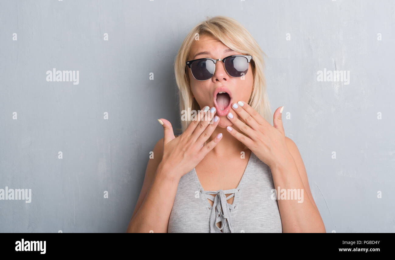 Adult caucasian woman over grunge grey wall wearing sunglasses scared ...