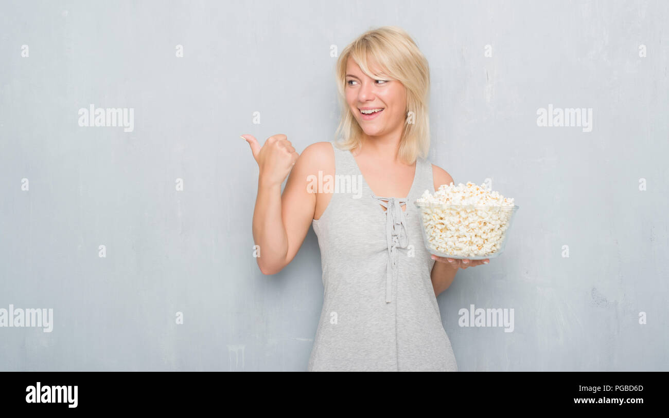 Adult caucasian woman over grunge grey wall eating pop corn pointing ...